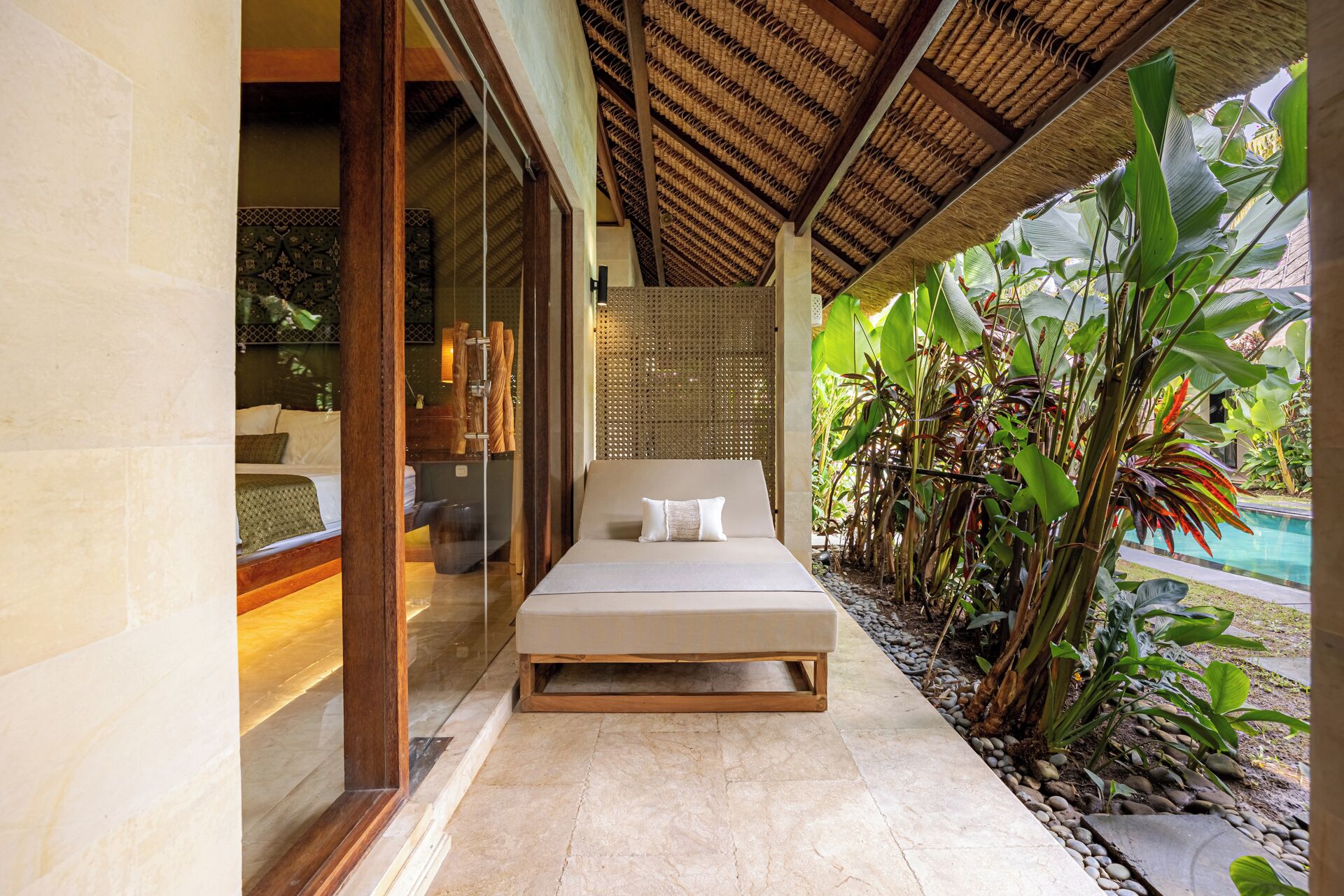 Quarto moderno com uma cama grande, porta de vidro que dá para um banheiro e vista para uma piscina e plantas tropicais.