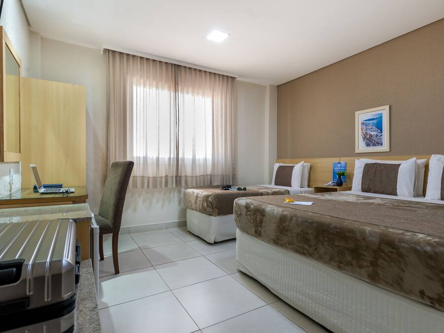 Quarto de hotel com duas camas, uma escrivaninha com um laptop, uma cadeira e uma janela grande com cortinas.