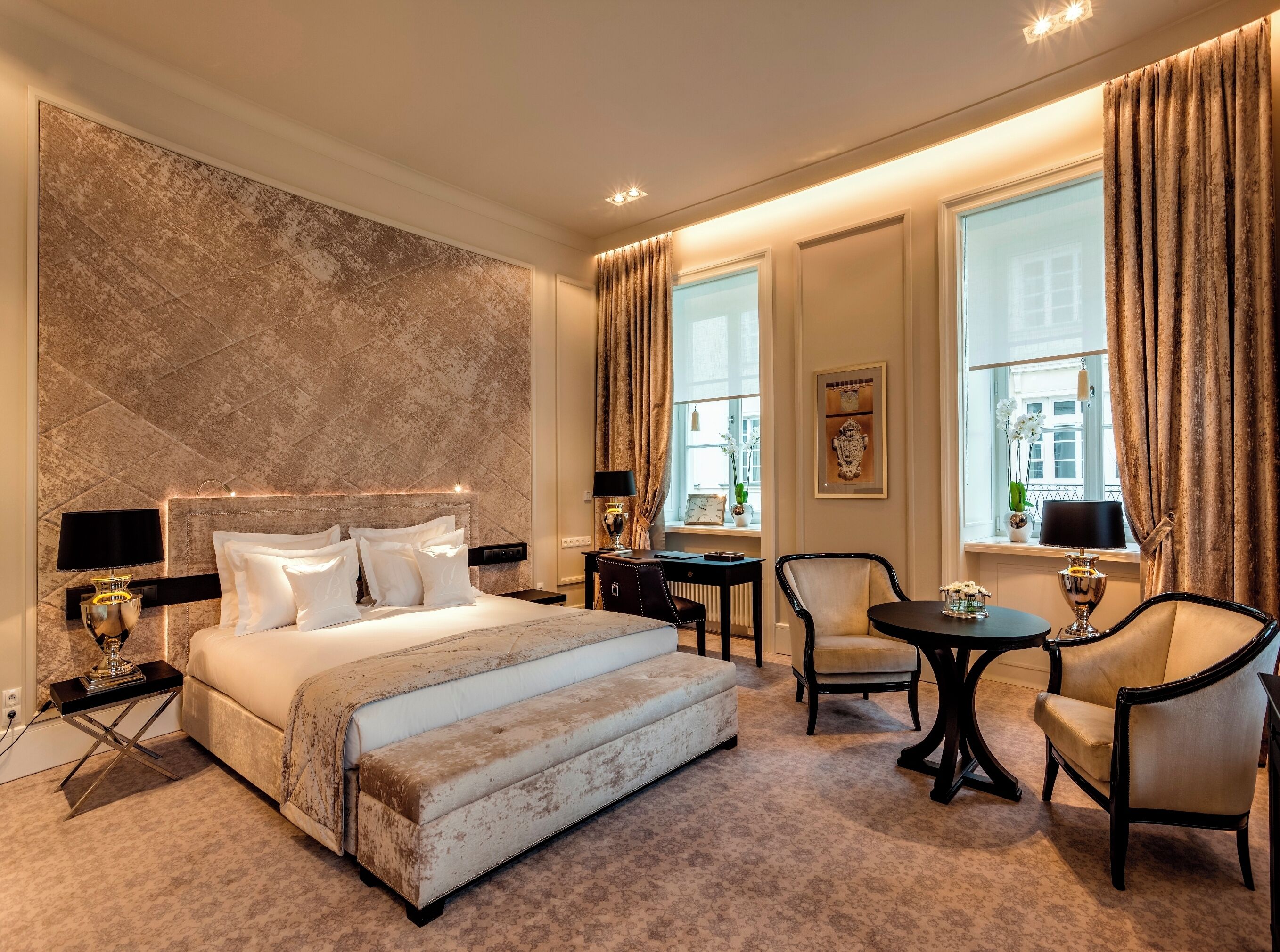 Quarto de hotel com uma cama grande, área de estar com mesa e cadeiras, janela com cortinas e um detalhe decorativo na parede.
