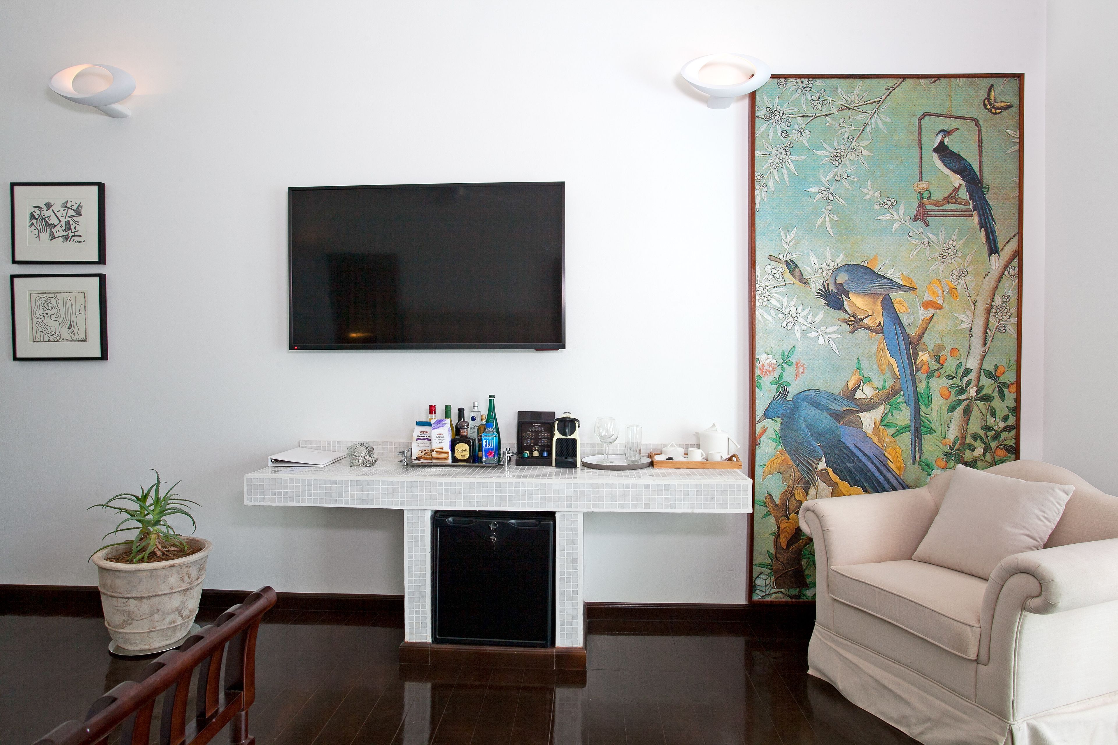 Uma sala de estar moderna com uma televisão de tela plana, uma mesa de centro revestida de azulejos brancos com diversos objetos, uma planta em vaso e um sofá.