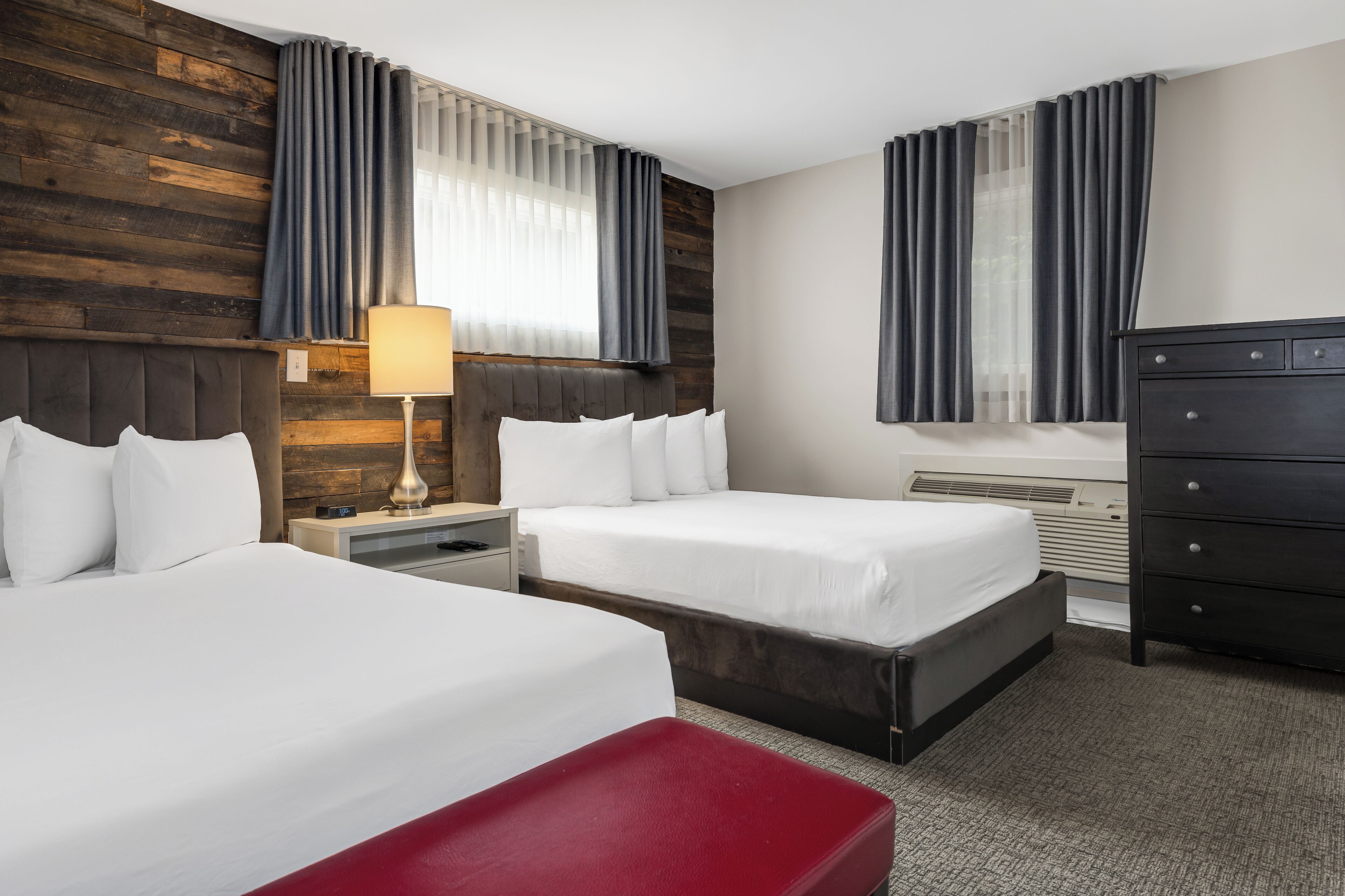 Quarto de hotel com duas camas, um banco vermelho, uma cômoda e um abajur.