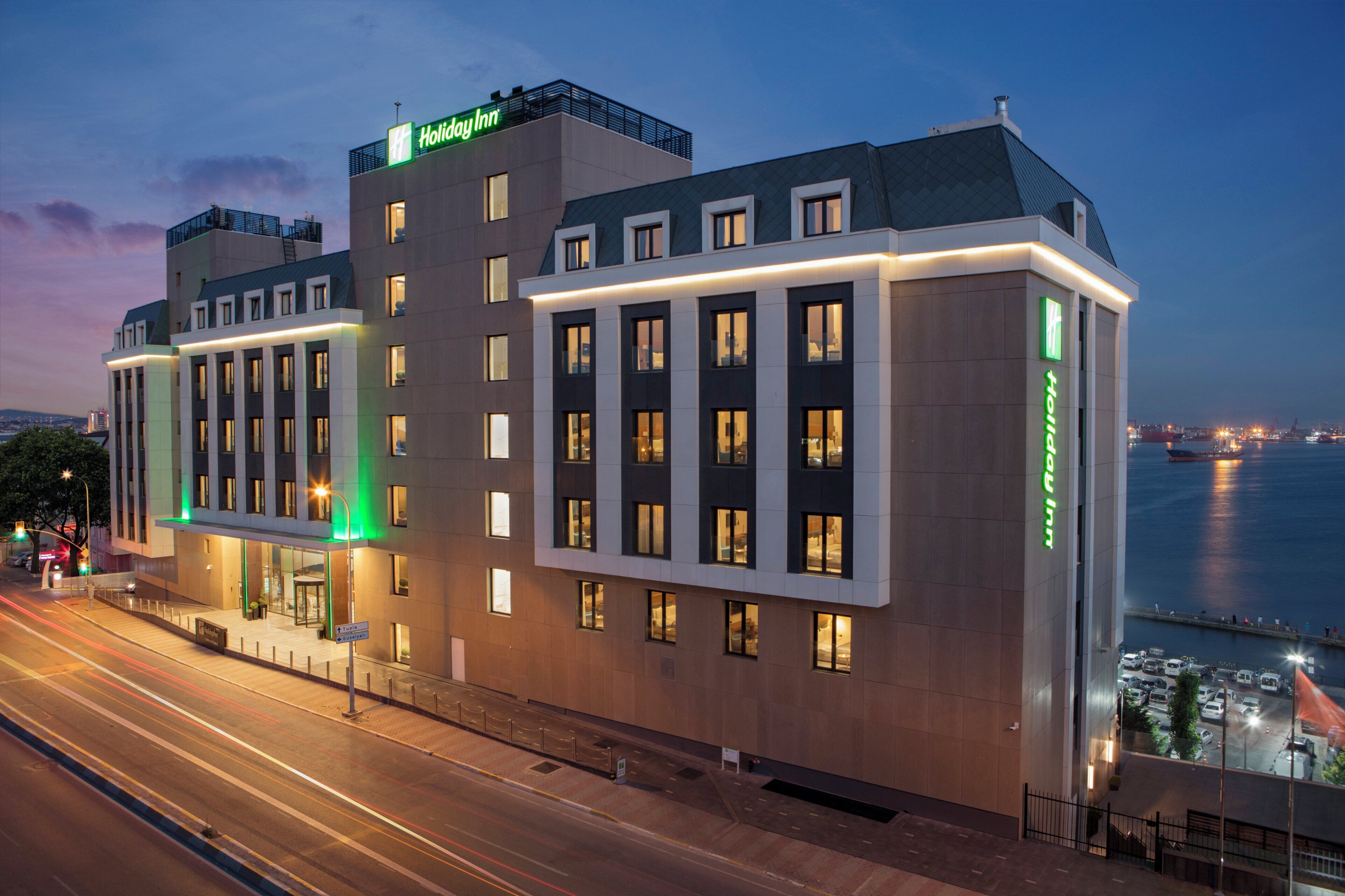 Um hotel moderno com uma placa Holiday Inn bem visível, vários andares e vista para a água e para o horizonte da cidade ao fundo.