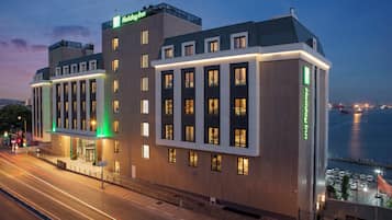 Um hotel moderno com uma placa Holiday Inn bem visível, vários andares e vista para a água e para o horizonte da cidade ao fundo.