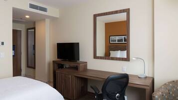 Quarto de hotel com uma cama, uma escrivaninha com televisão, uma cadeira e um espelho grande.