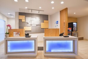Uma área de recepção de hotel com dois balcões, uma planta e um refrigerador abastecido com bebidas.