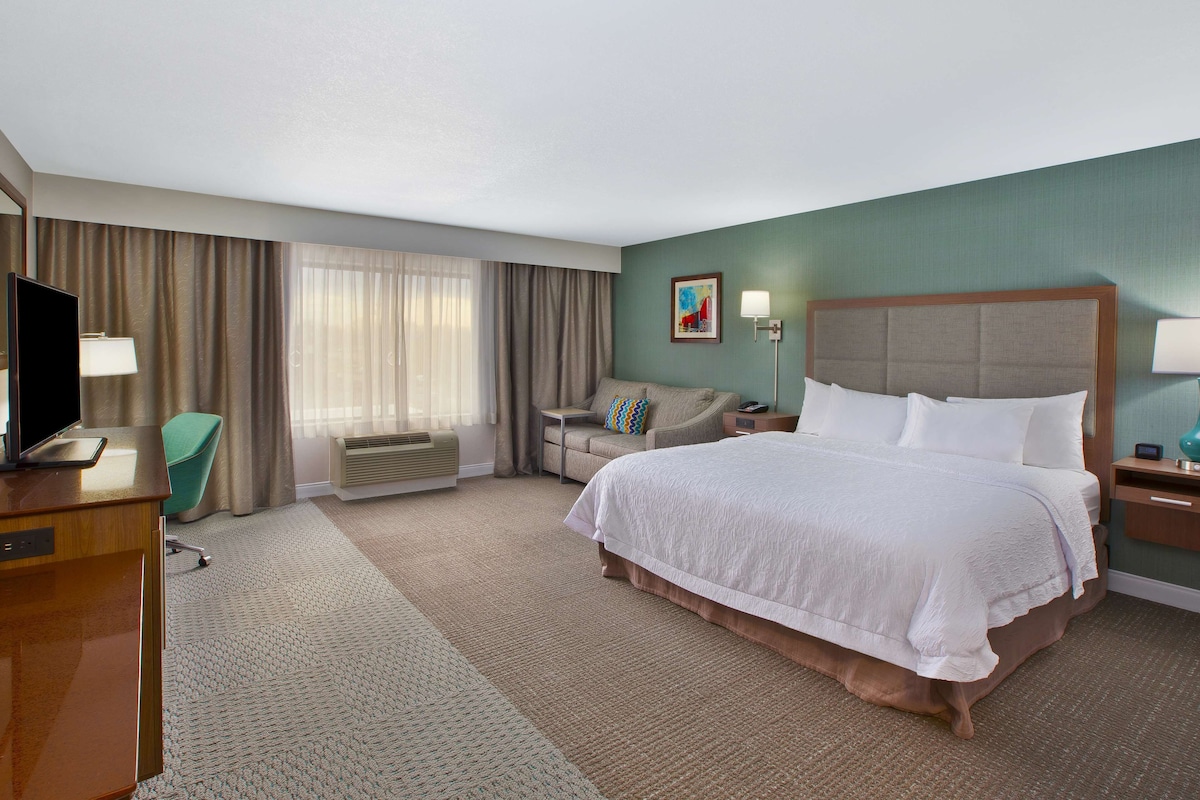 Quarto de hotel com cama, escrivaninha, cadeira e televisão.