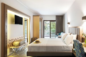 Quarto de hotel com uma cama grande, televisão na parede, mala num banco e varanda com vista para árvores.