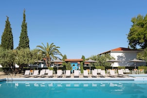 Piscina com espreguiçadeiras e guarda-sóis, cercada por árvores e edifícios.