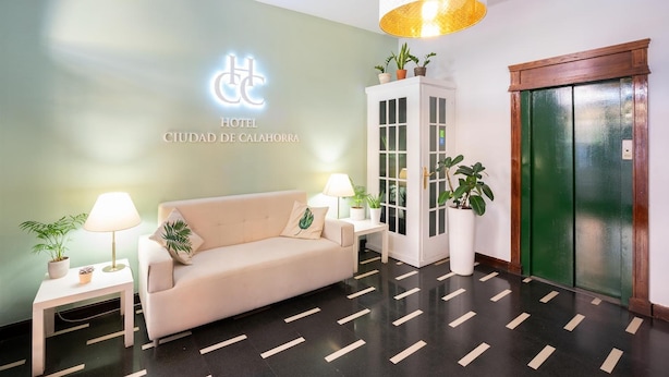 Um saguão de hotel com um sofá, duas luminárias, um vaso de planta, um armário e uma porta de elevador verde.