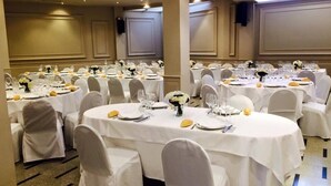 Salão de banquetes com mesas redondas postas para refeição, toalhas brancas e cadeiras com capas brancas.