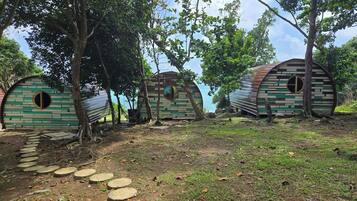 Três cabanas no estilo de casas na árvore com janelas circulares, rodeadas por árvores e um caminho de pedra.