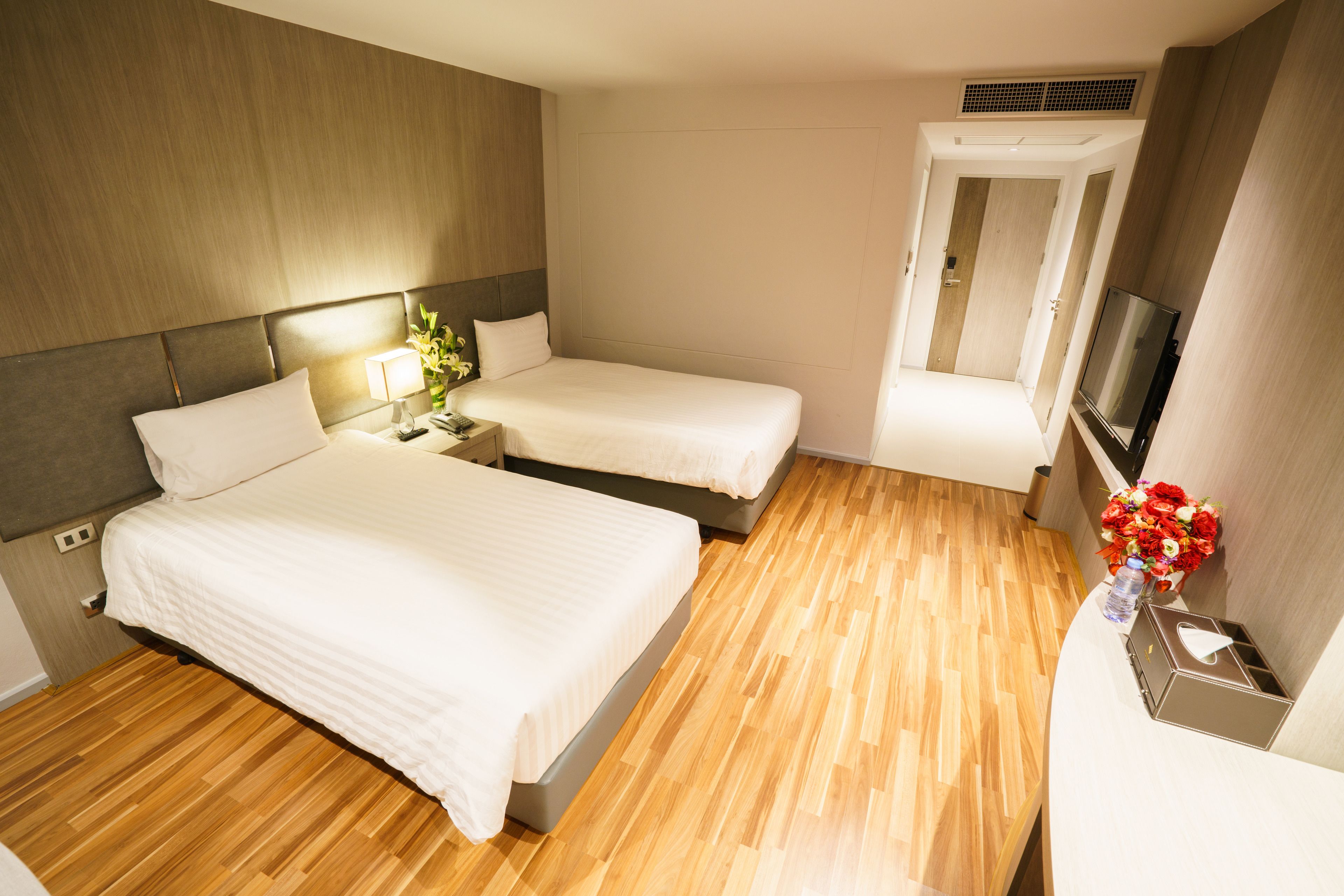 Quarto de hotel com duas camas, televisão, escrivaninha com um vaso de flores e piso de madeira.