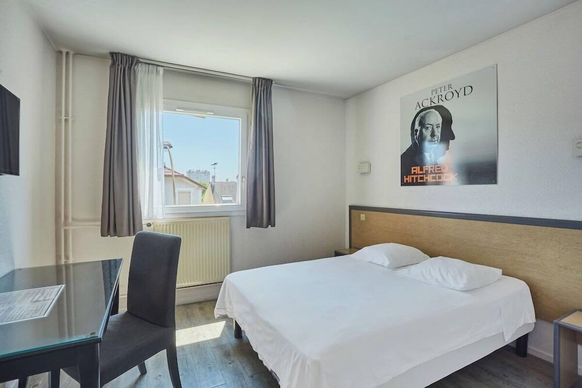 Quarto de hotel com cama, escrivaninha, cadeira, janela com cortinas e um cartaz na parede.