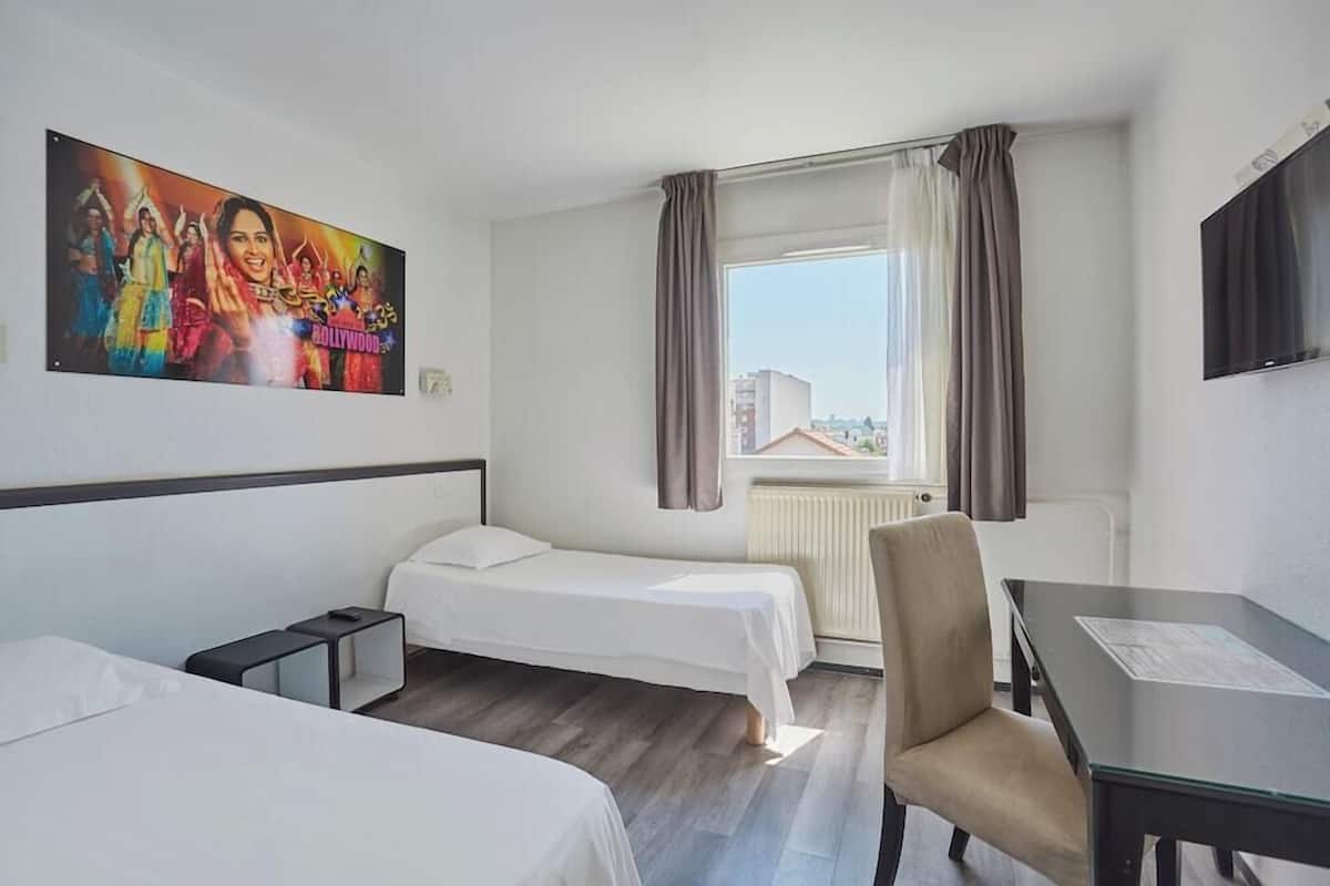 Quarto de hotel com duas camas, uma escrivaninha, uma cadeira, janela com cortinas, radiador e televisão presa na parede.