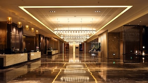 Um hall amplo com piso de mármore refletivo, um lustre central e um balcão de recepção à esquerda.