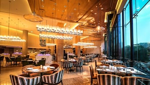 Um restaurante espaçoso com várias áreas de refeição, grandes janelas com vista para a cidade e lustres modernos.