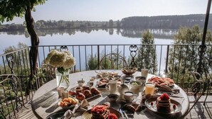 Mesa redonda para refeições com diversos itens de café da manhã e um vaso de flores, em uma varanda com vista para um lago.