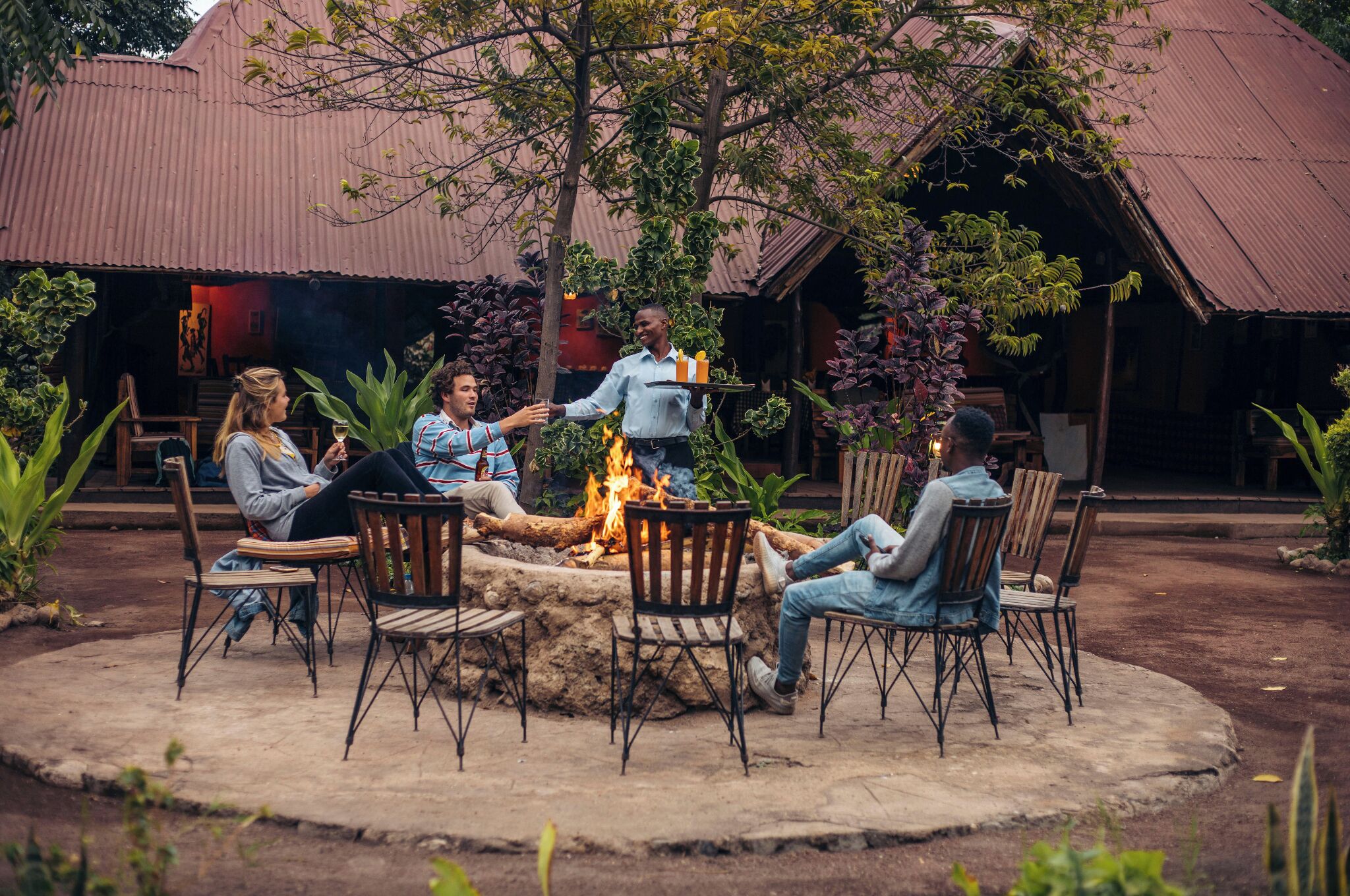 Um grupo de pessoas sentadas ao redor de uma fogueira em ambiente externo, com edifícios rústicos ao fundo.