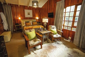 Um quarto aconchegante com piso de madeira, uma cama com cabeceira, uma mesa com cadeiras, uma televisão fixada na parede e uma janela com cortinas.