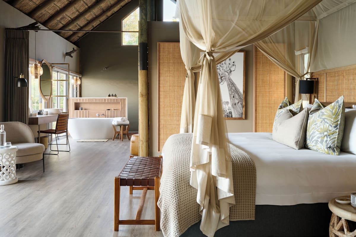 Quarto com cama de dossel, área de estar com poltrona e mesa, um espelho grande e piso de madeira.