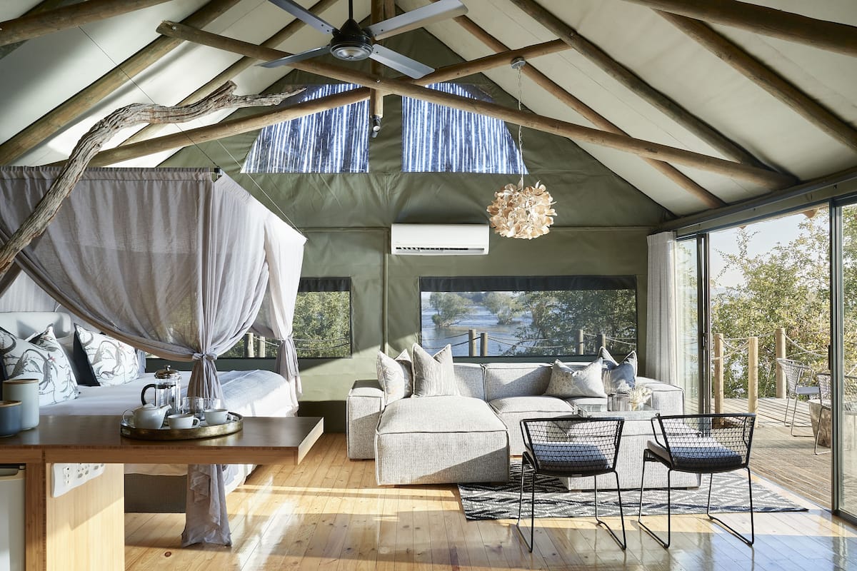 Quarto espaçoso com cama grande, sofá e área de refeições. Há um ventilador de teto, luminária pendente e vista para o exterior por grandes janelas.