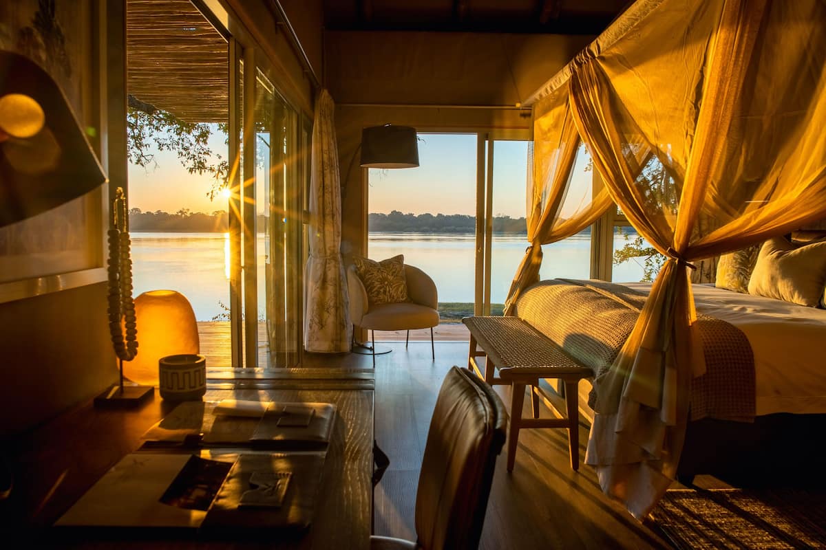 Quarto com cama, escrivaninha e cadeira, com vista para a água e árvores.