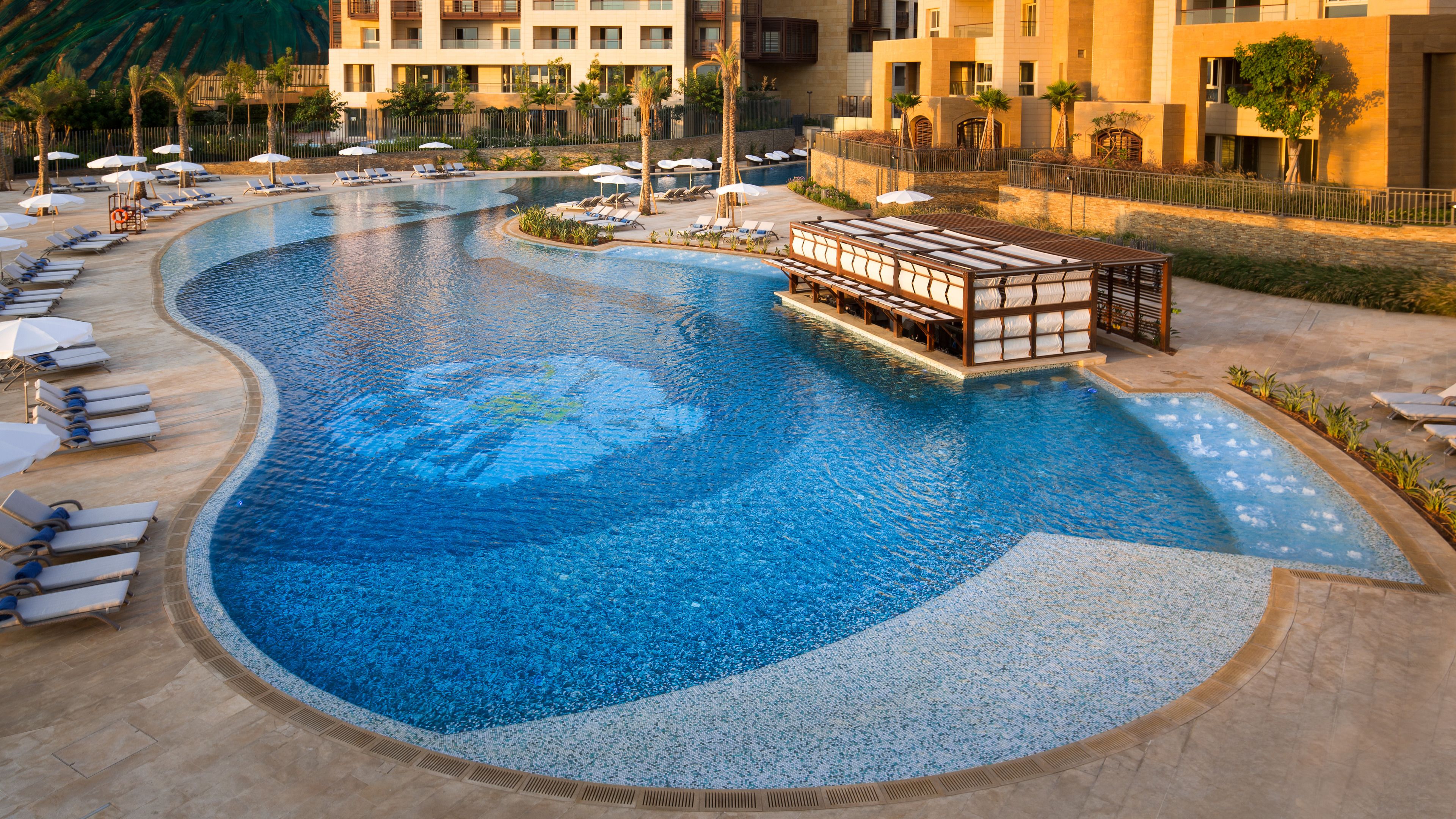 Uma grande piscina com espreguiçadeiras e guarda-sóis, rodeada por um edifício com varandas.
