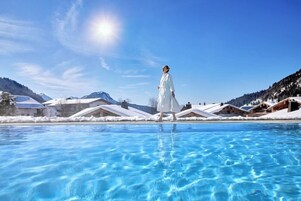 Uma pessoa vestida com um robe branco caminhando sobre uma piscina azul cristalina, rodeada por edifícios cobertos de neve e montanhas.