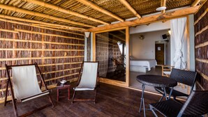 Quarto com teto de bambu trançado, paredes de madeira e uma porta deslizante com espelho que dá para a cama. Há duas cadeiras de lona e uma mesa pequena.