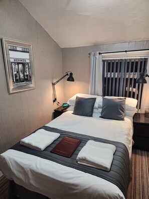 Quarto de hotel com cama arrumada, quadro com a imagem de Hermes na parede, mesa de cabeceira com abajur e janela com persianas.