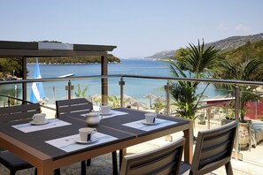 Terraço com mesa posta para dois, com vista para a costa, guarda-sóis e plantas.