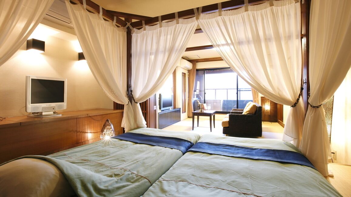 Uma cama de casal com roupa de cama branca e azul, uma televisão à esquerda e vista para uma varanda com cadeira e mesa.
