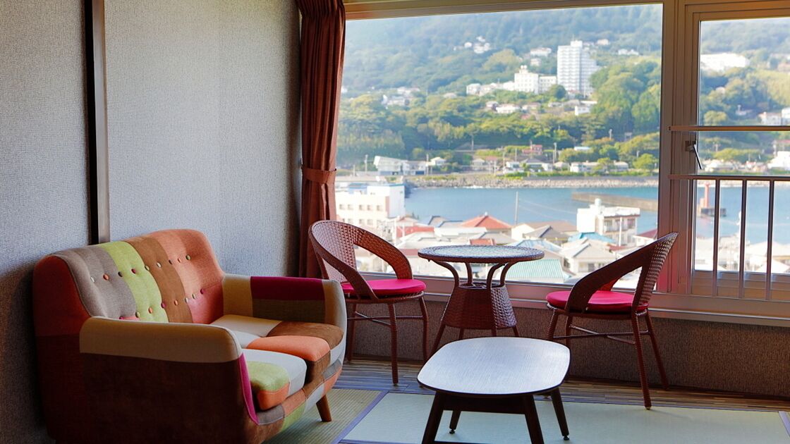 Quarto com sofá, duas cadeiras, mesa pequena e grande janela com vista para uma cidade e colinas.