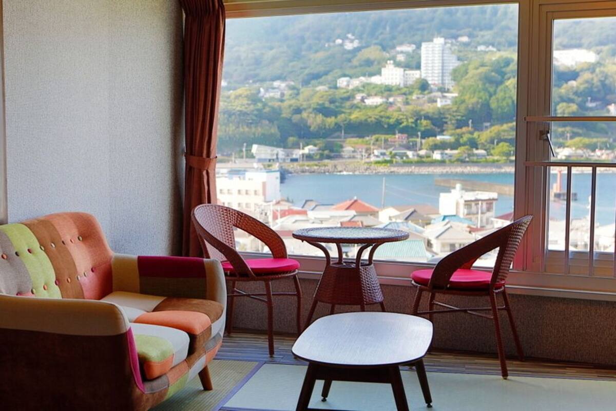 Quarto com sofá, duas cadeiras, mesa pequena e grande janela com vista para uma cidade e colinas.