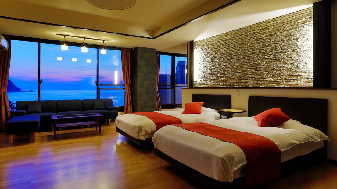 Quarto de hotel com duas camas, um sofá, um banco e uma janela grande com vista para o mar.
