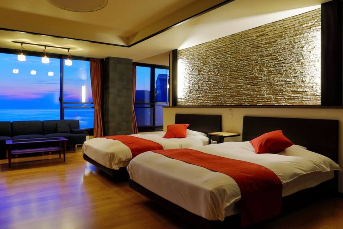 Quarto de hotel com duas camas, um sofá, um banco e uma janela grande com vista para o mar.