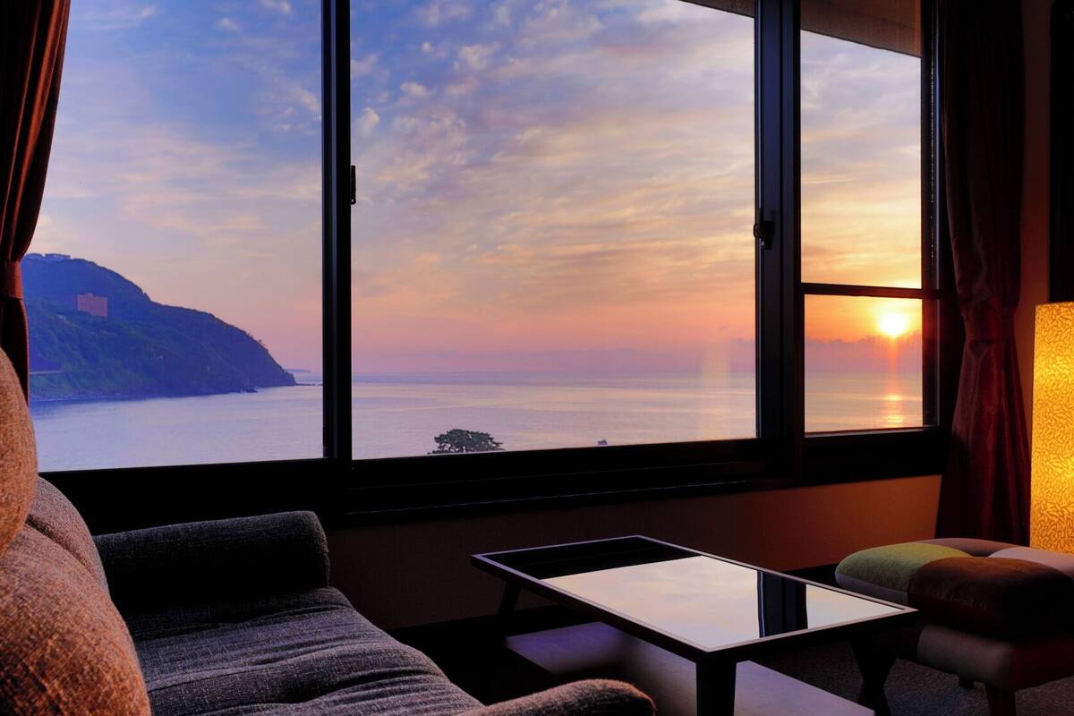 Quarto com uma grande janela com vista para a água e o pôr do sol, um sofá, uma mesinha e uma luminária.