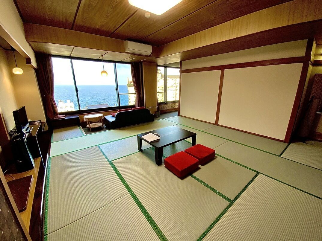 Quarto com piso de tatame, mesa de tatame, um banco vermelho, uma televisão e uma janela grande com vista para o mar.