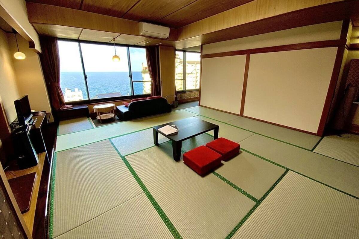 Quarto com piso de tatame, mesa de tatame, um banco vermelho, uma televisão e uma janela grande com vista para o mar.