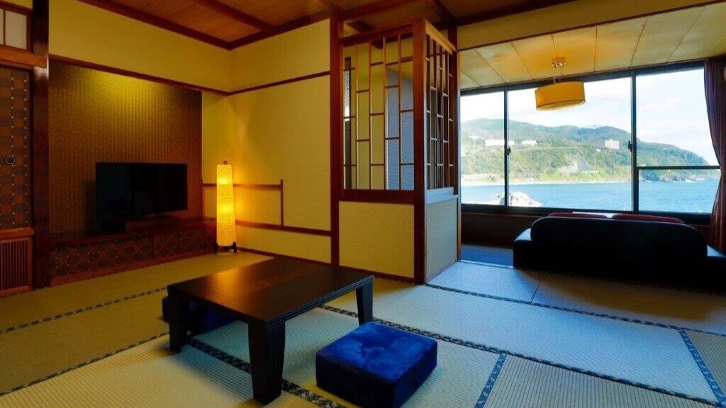 Um quarto japonês tradicional com tatames, uma mesa baixa, uma televisão e vista para o mar.