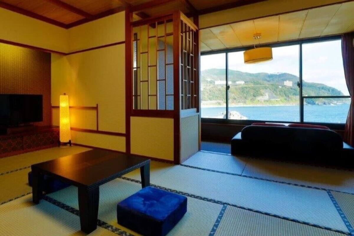 Um quarto japonês tradicional com tatames, uma mesa baixa, uma televisão e vista para o mar.