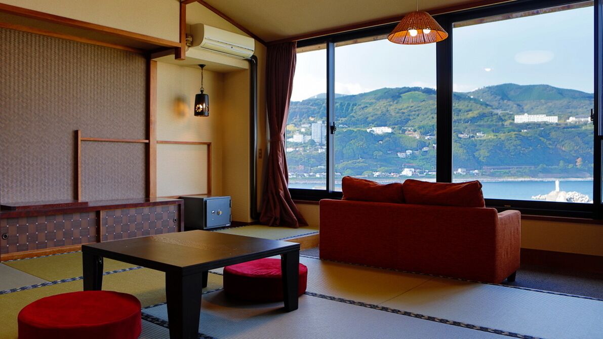 Quarto com piso de tatame, uma mesa baixa, um sofá vermelho e uma janela grande com vista para colinas e um corpo d'água.