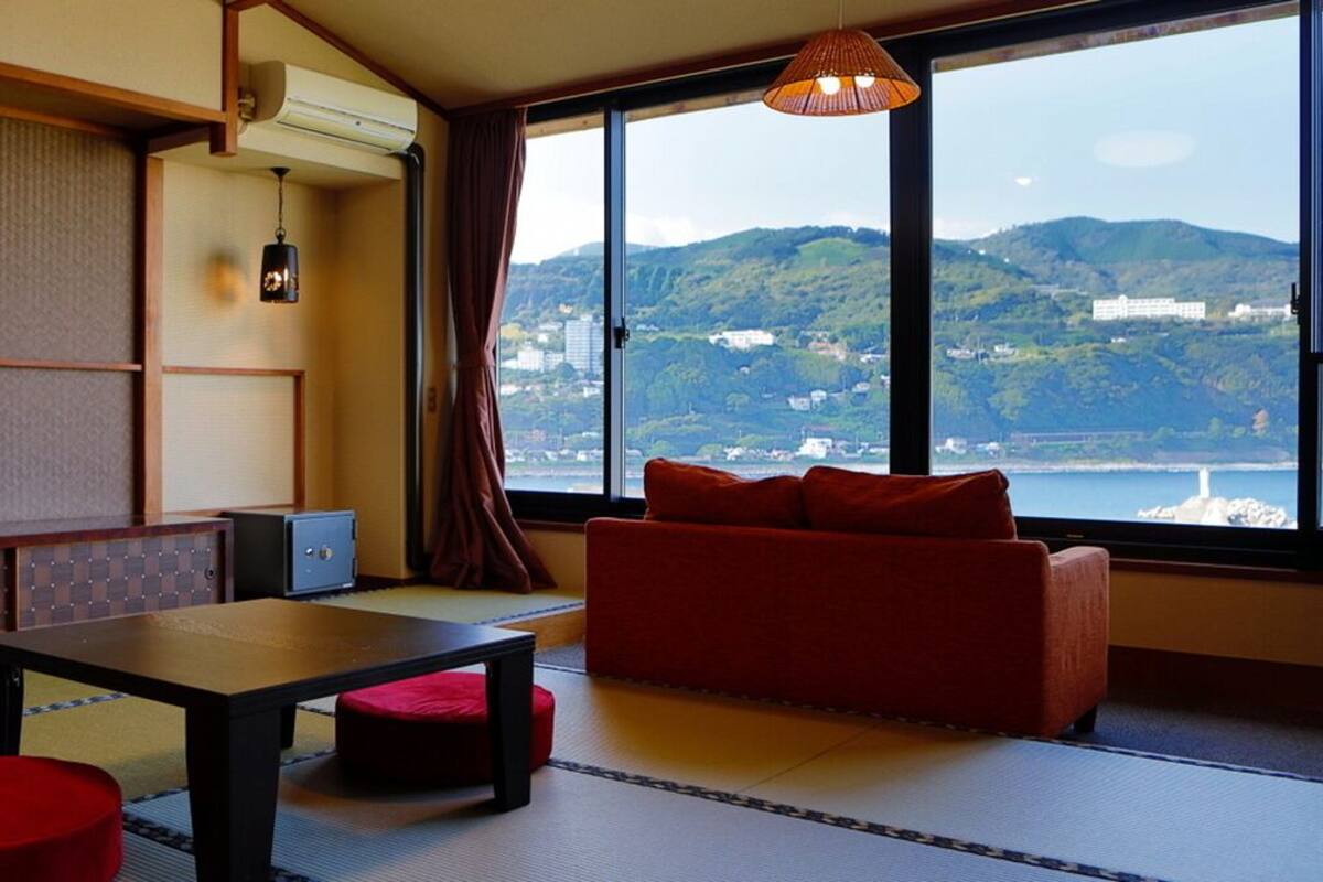 Quarto com piso de tatame, uma mesa baixa, um sofá vermelho e uma janela grande com vista para colinas e um corpo d'água.
