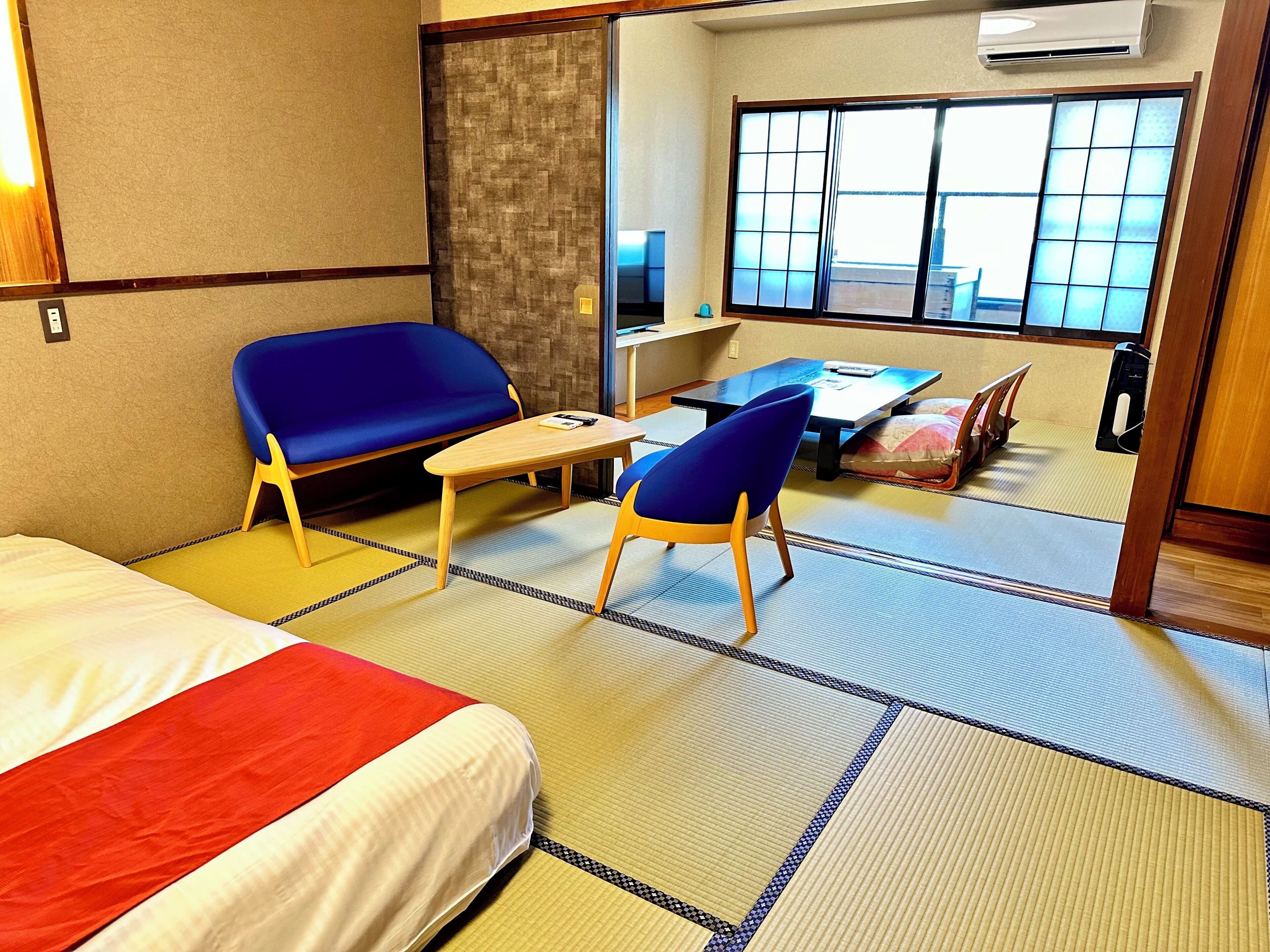 Quarto de hotel com piso de tatame, cama com manta vermelha, uma pequena mesa, duas cadeiras azuis e uma porta deslizante para uma varanda.