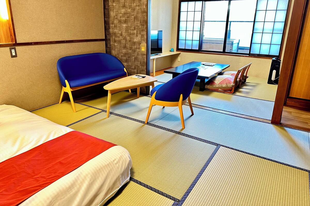 Quarto de hotel com piso de tatame, cama com manta vermelha, uma pequena mesa, duas cadeiras azuis e uma porta deslizante para uma varanda.