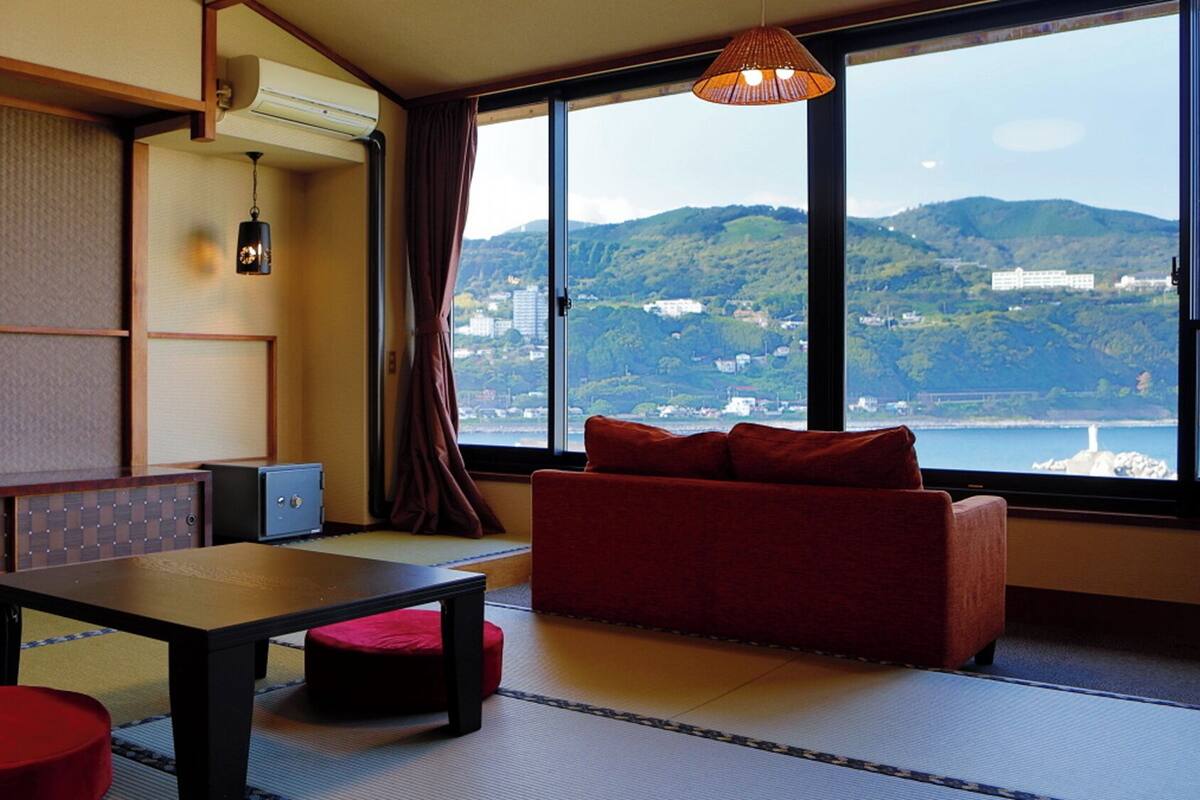 Quarto com piso de tatame, uma mesinha baixa, um sofá e uma janela grande com vista para colinas e edifícios.