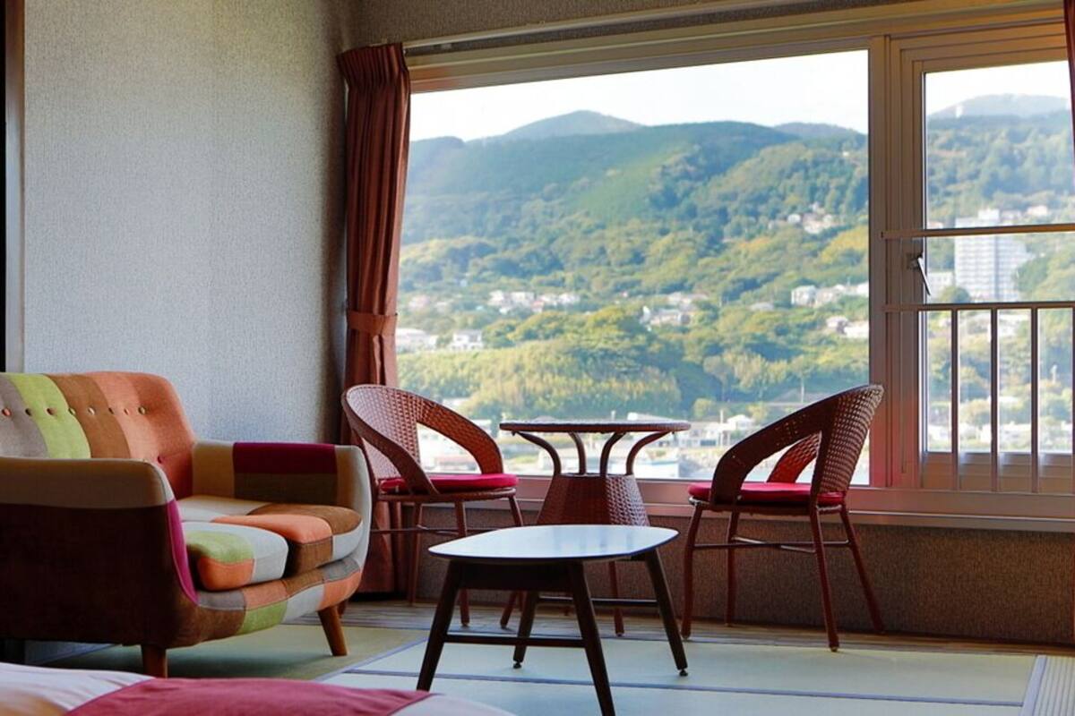 Quarto com sofá, duas cadeiras, mesa pequena e janela com vista para uma paisagem montanhosa.