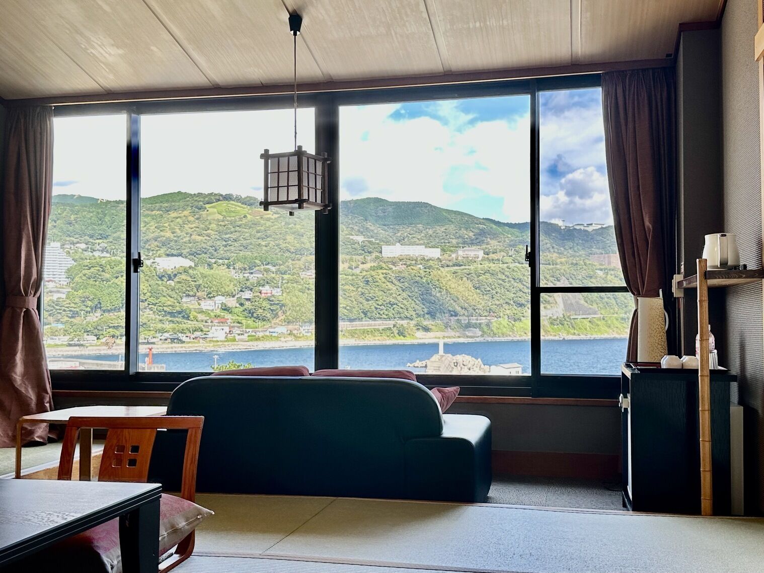 Quarto com uma janela grande que oferece vista para uma paisagem acidentada e um corpo d'água. Há um sofá, uma mesa pequena e um lustre.