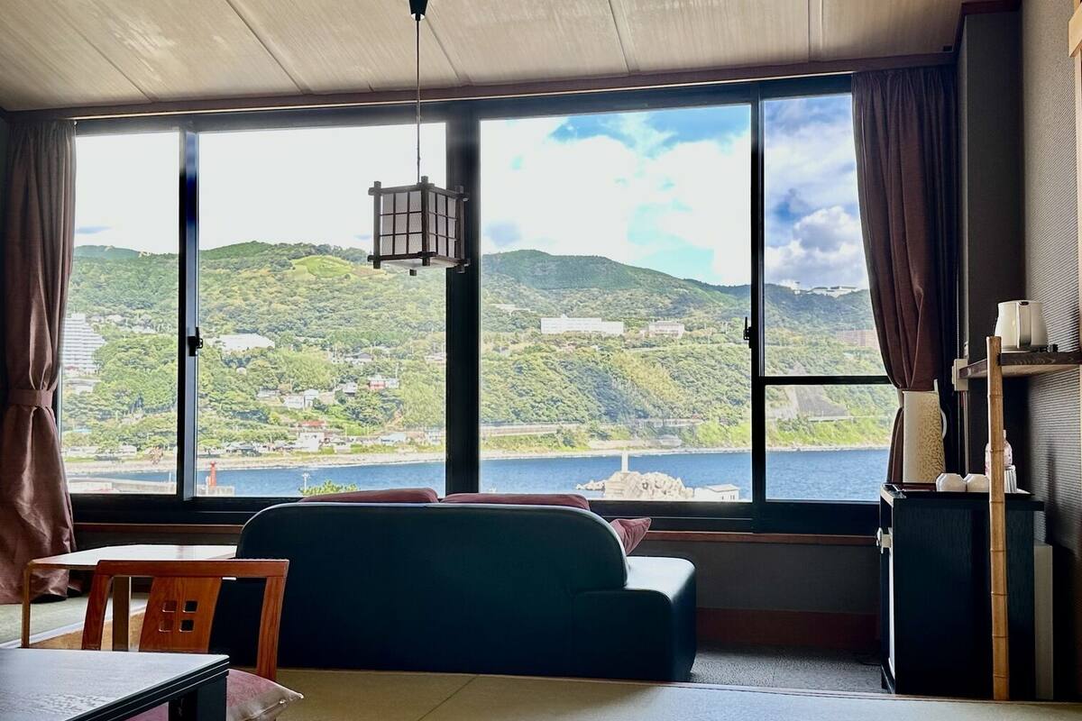 Quarto com uma janela grande que oferece vista para uma paisagem acidentada e um corpo d'água. Há um sofá, uma mesa pequena e um lustre.