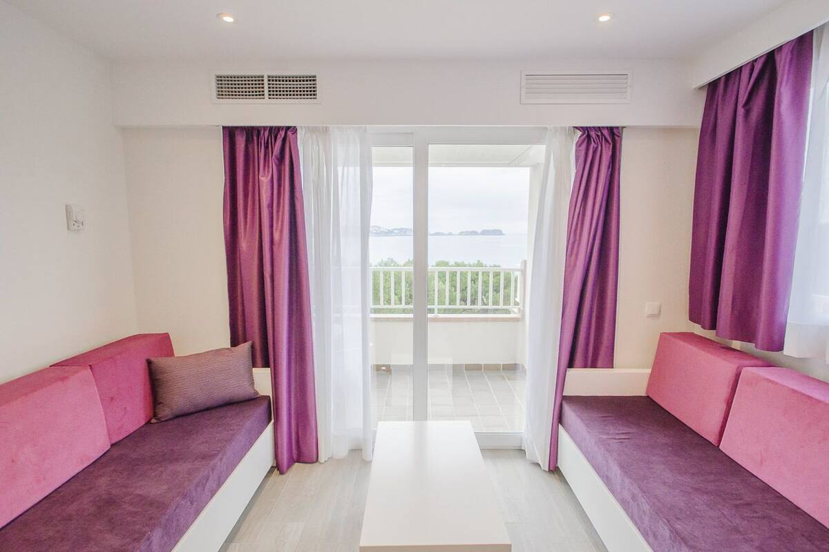Quarto com dois sofás cor-de-rosa, uma mesa branca e uma varanda com cortinas roxas.